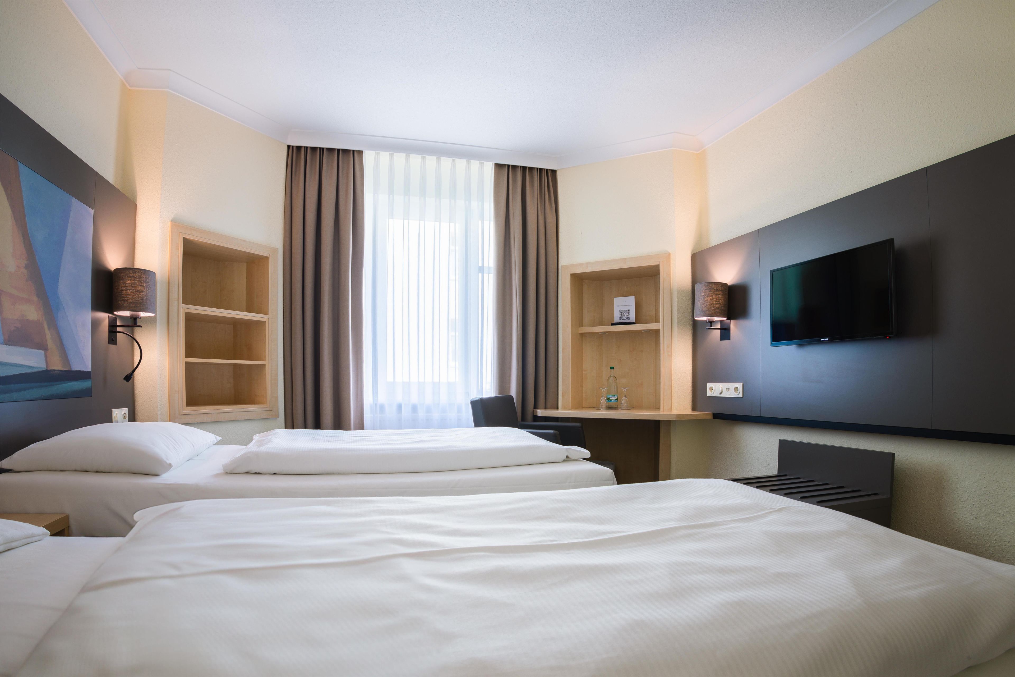 Quarto de hotel com duas camas, uma escrivaninha, uma cadeira, uma televisão e uma janela com cortinas.