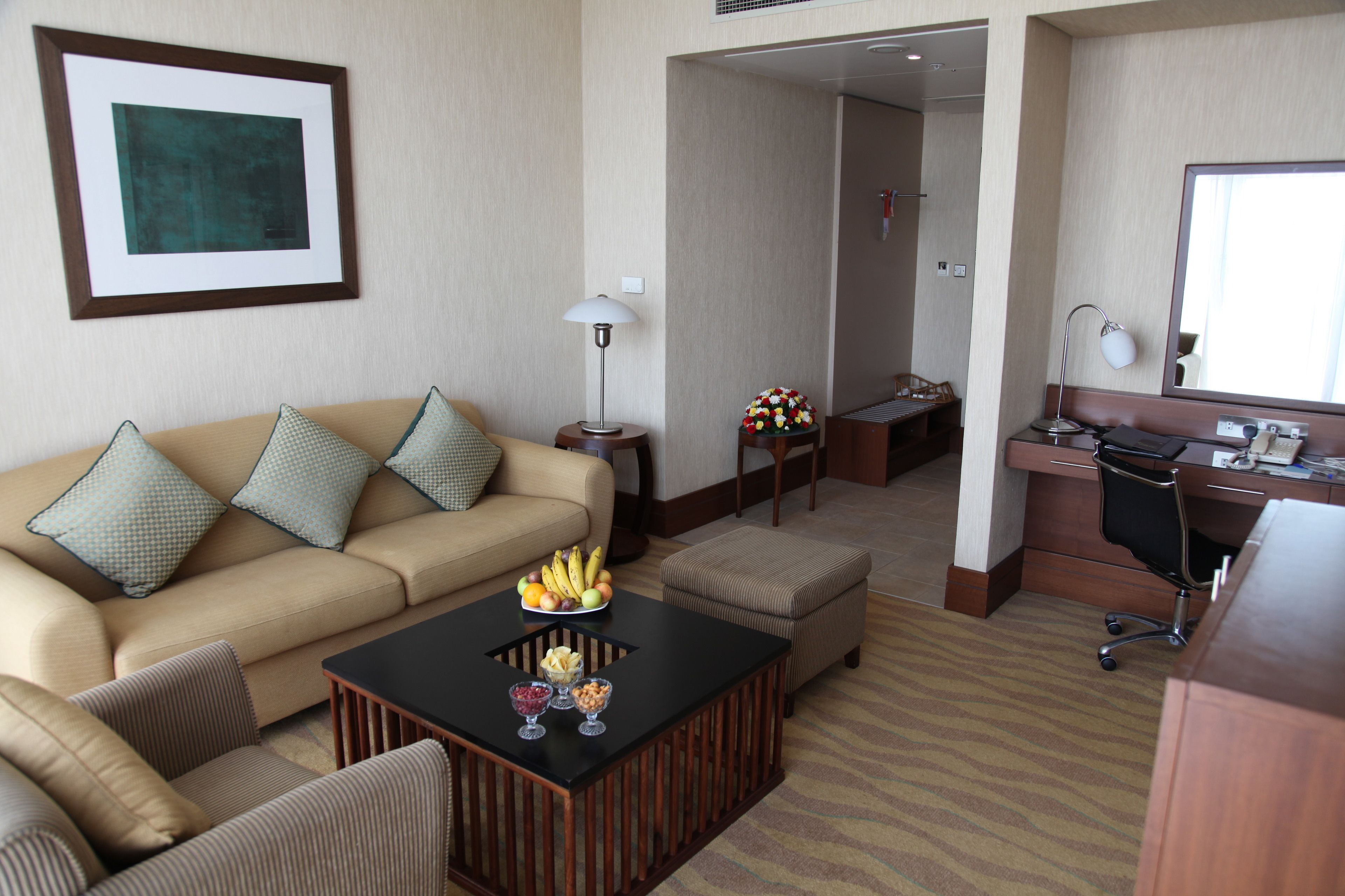 Quarto de hotel com sofá, mesa de centro com frutas e tigelas, escrivaninha com cadeira e um espelho grande.