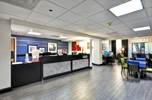Uma área de recepção de hotel com balcão, parede azul com placa de boas-vindas, área de estar com cadeiras e mesas, e design de interiores moderno.