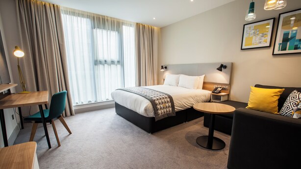 Quarto de hotel moderno com uma cama, uma escrivaninha, uma cadeira, um sofá e uma mesinha.