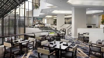 Um lobby de hotel moderno com várias áreas de refeições, uma escadaria e vista para a paisagem urbana.