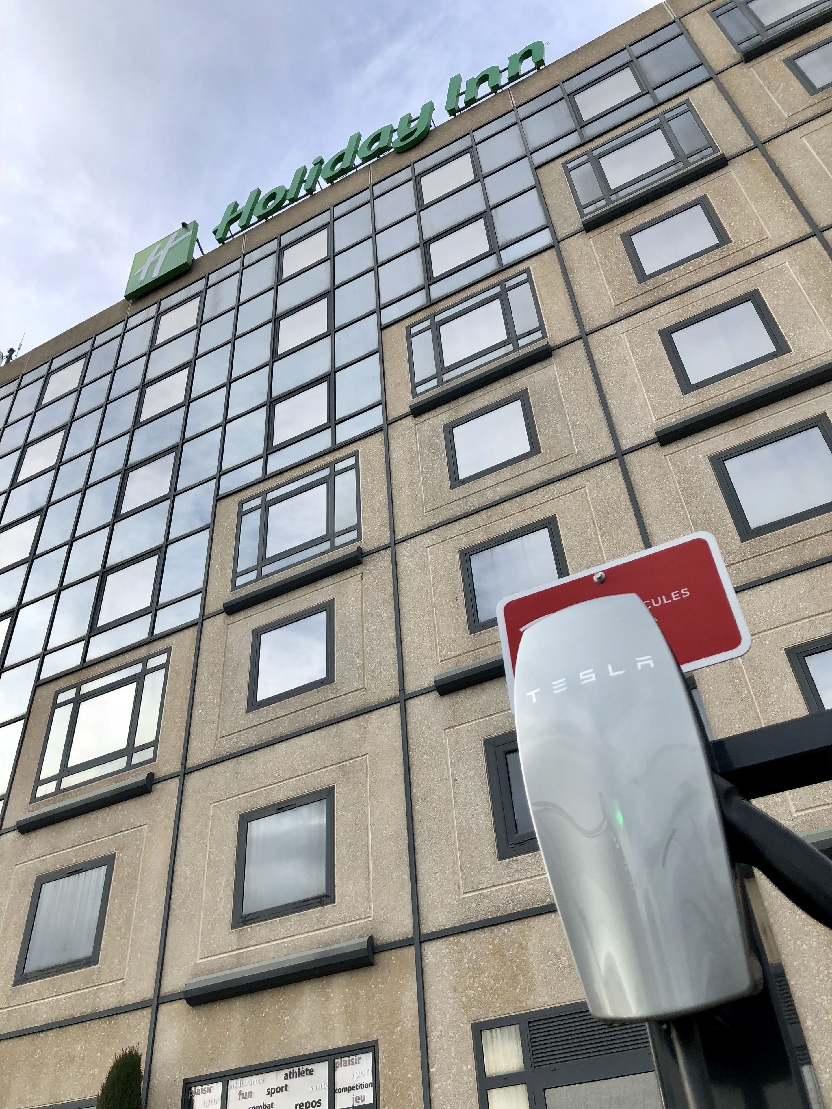 Edifício Holiday Inn com fachada moderna, grandes janelas e uma estação de carregamento Tesla em frente.