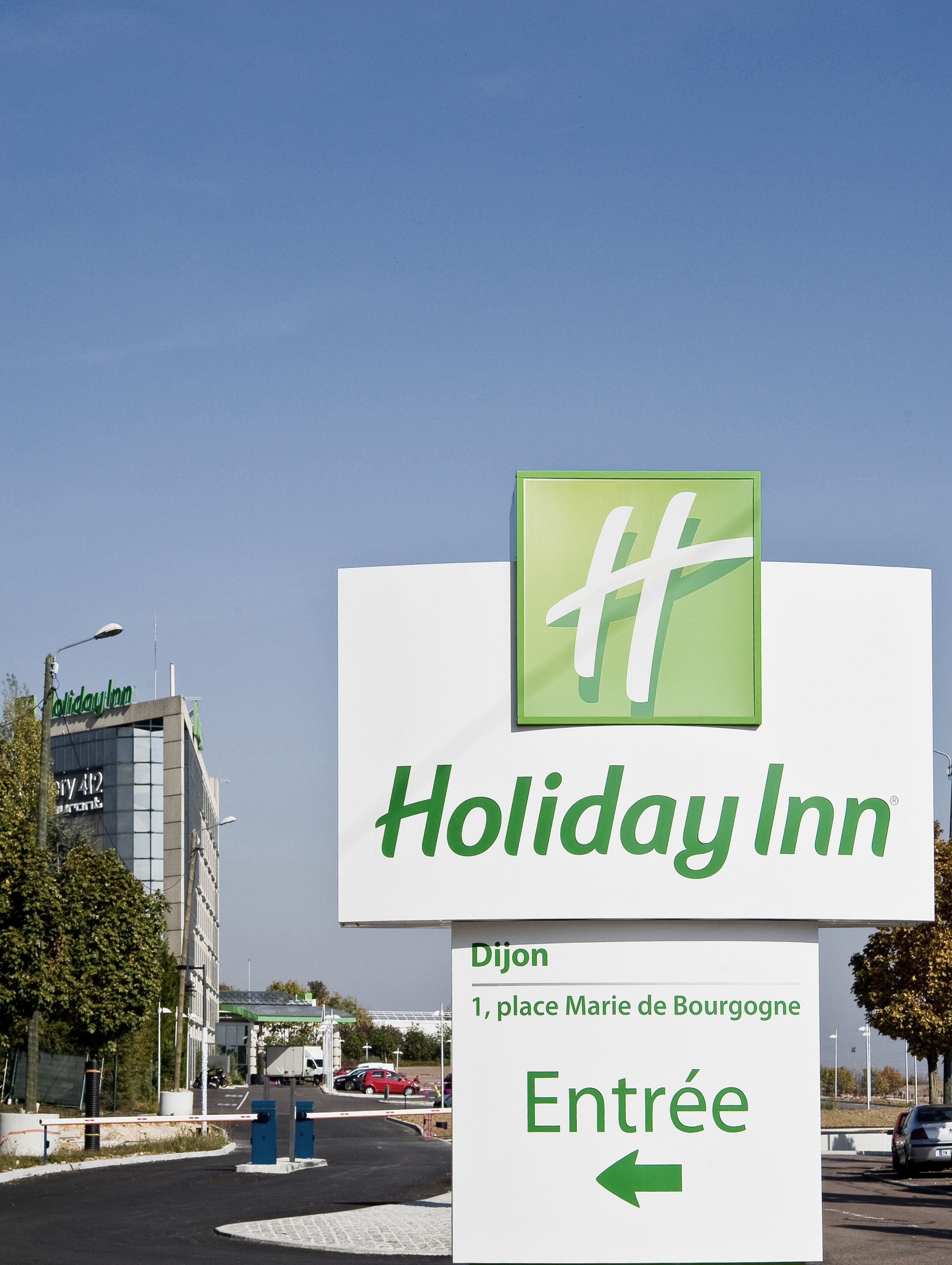 Placa de um Holiday Inn com logotipo verde e o nome 'Holiday Inn' em letras grandes. Abaixo, uma placa de direção indica 'Dijon 1, place Marie de Bourgogne' e uma seta apontando para a esquerda para 'Entrée'.