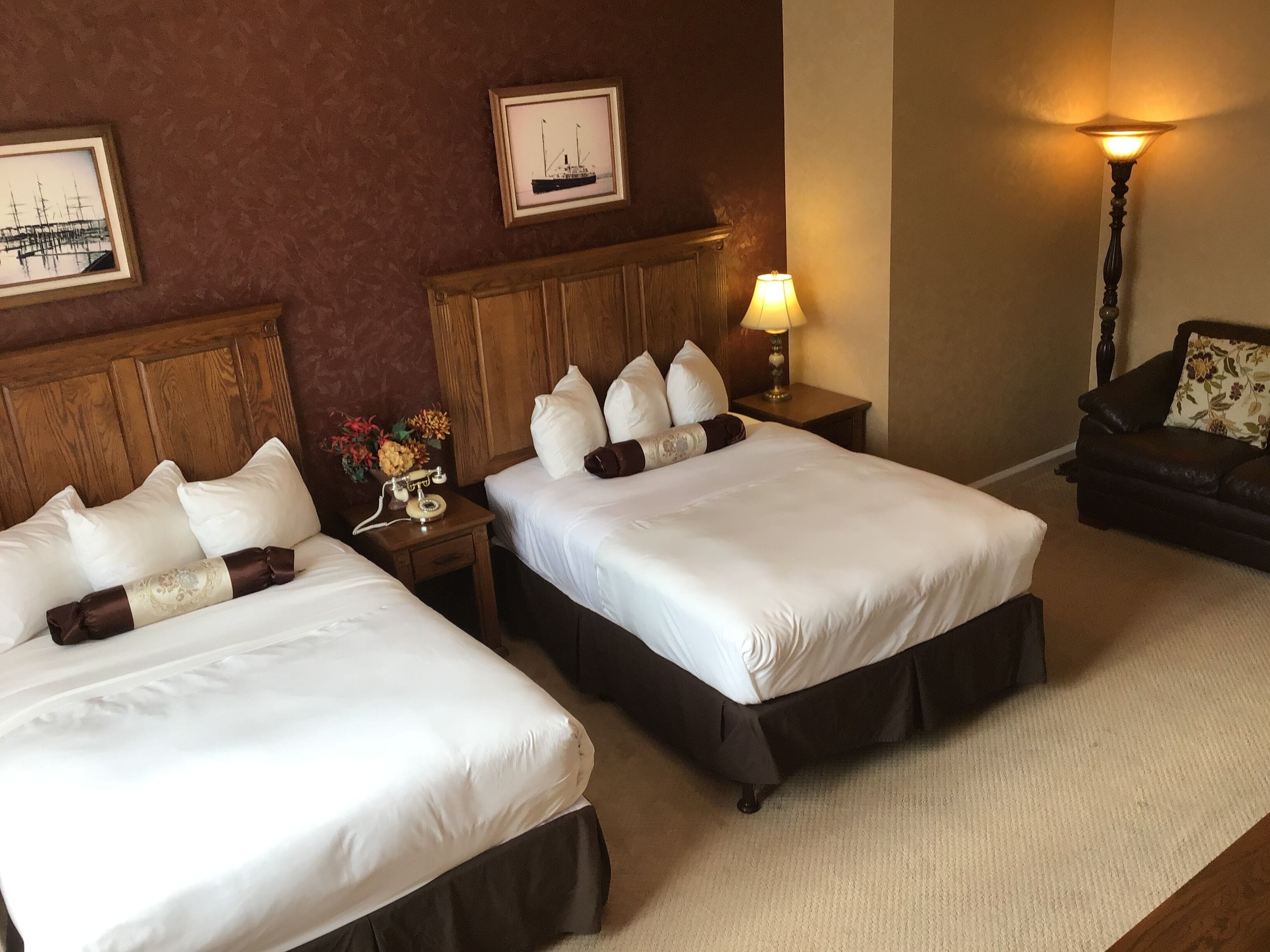 Quarto de hotel com duas camas, cabeceira de madeira, mesa de cabeceira com abajur, quadro emoldurado na parede e um sofá.
