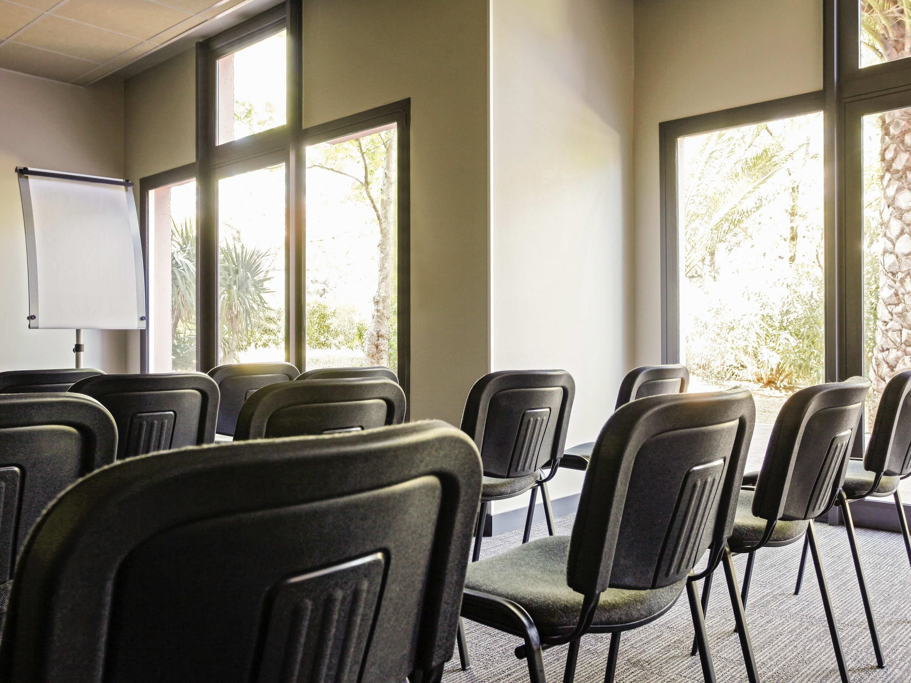 Uma sala de conferências com fileiras de cadeiras, um quadro branco e grandes janelas com vista para árvores.