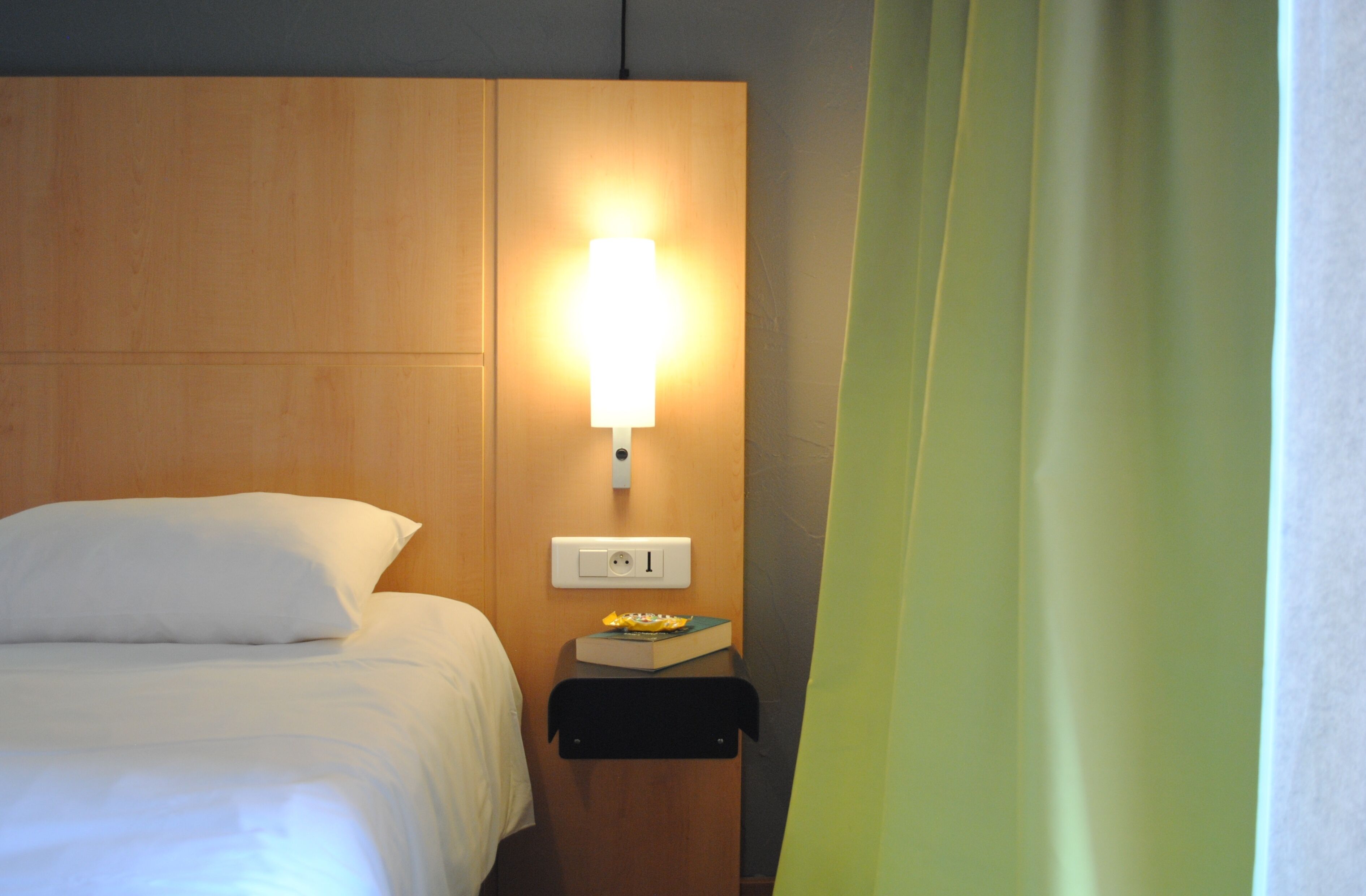 Quarto de hotel com uma cama, mesa de cabeceira com abajur, um livro e uma tomada.