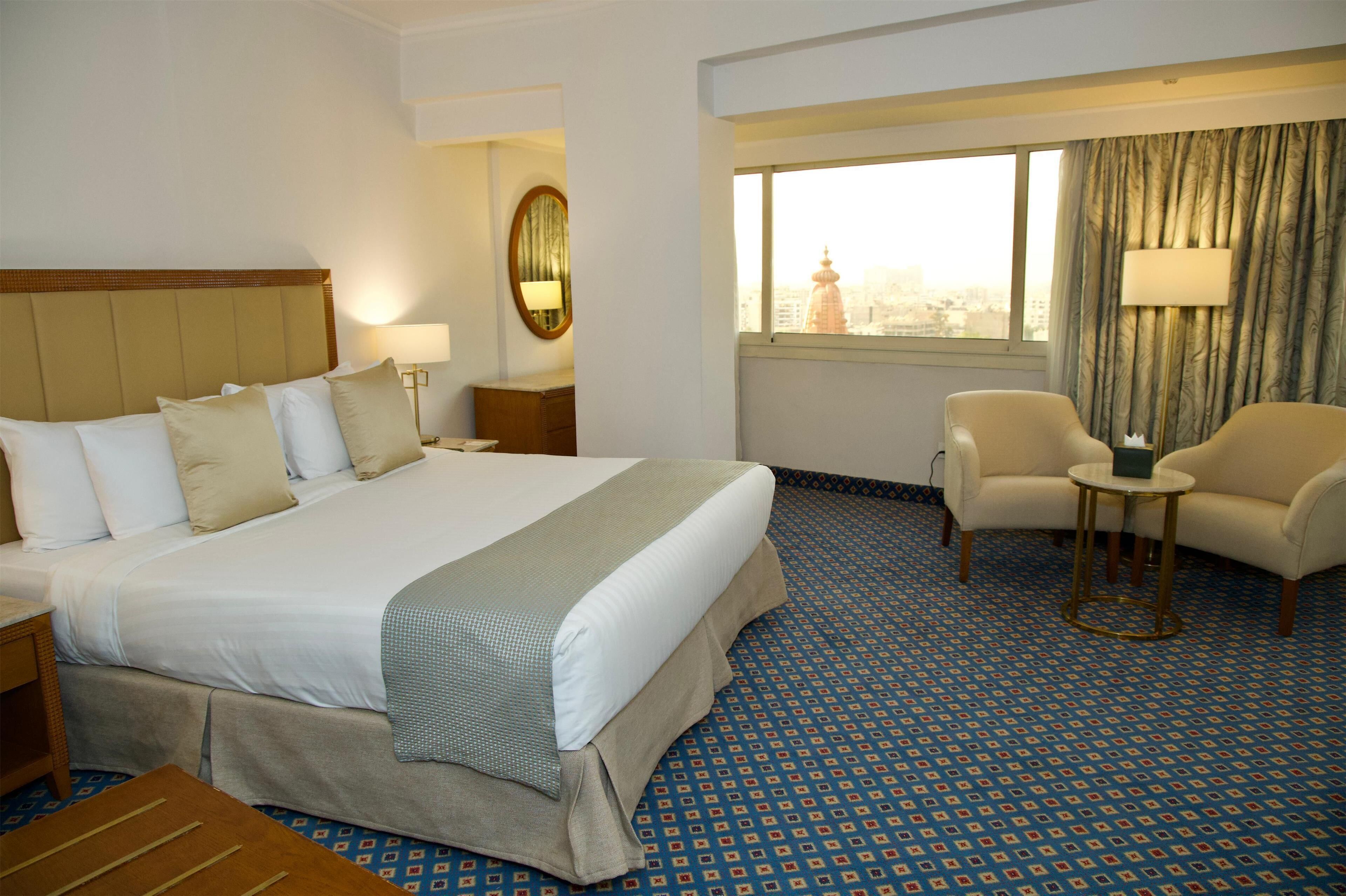 Quarto de hotel com uma cama grande, duas poltronas, uma mesa pequena, uma mesa de cabeceira e uma janela com cortinas.