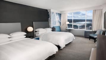 Quarto de hotel com duas camas, uma escrivaninha, uma cadeira e uma janela grande com vista para um aeroporto.