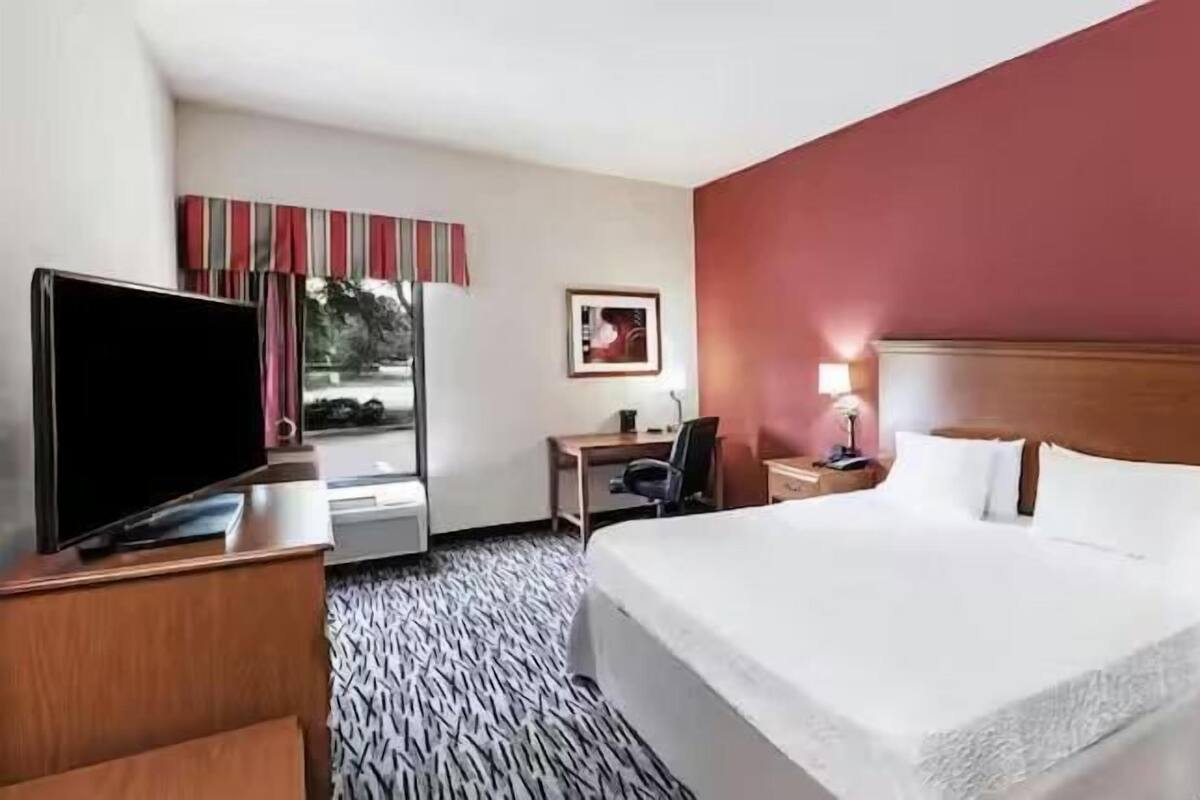 Quarto de hotel com cama, escrivaninha, cadeira, televisão e janela com cortinas.