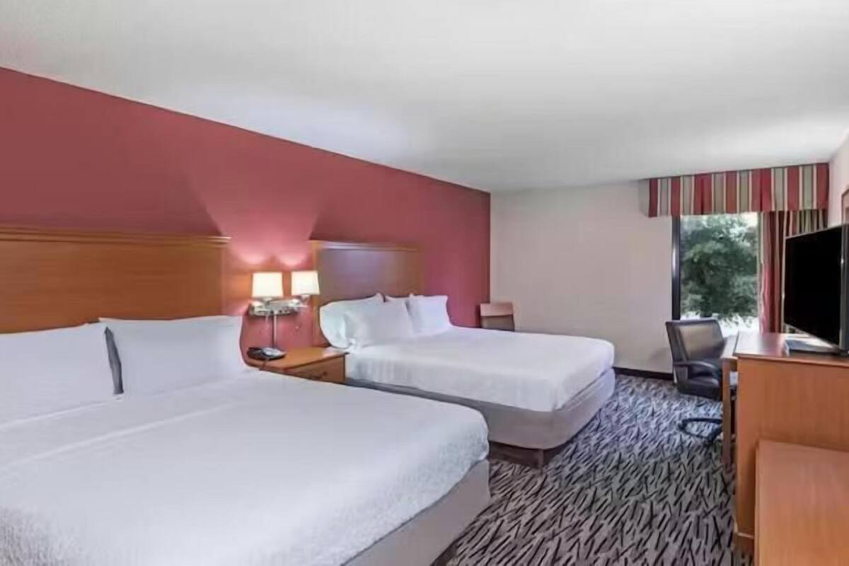 Quarto de hotel com duas camas, uma mesa, uma cadeira, uma TV e uma janela com cortinas.