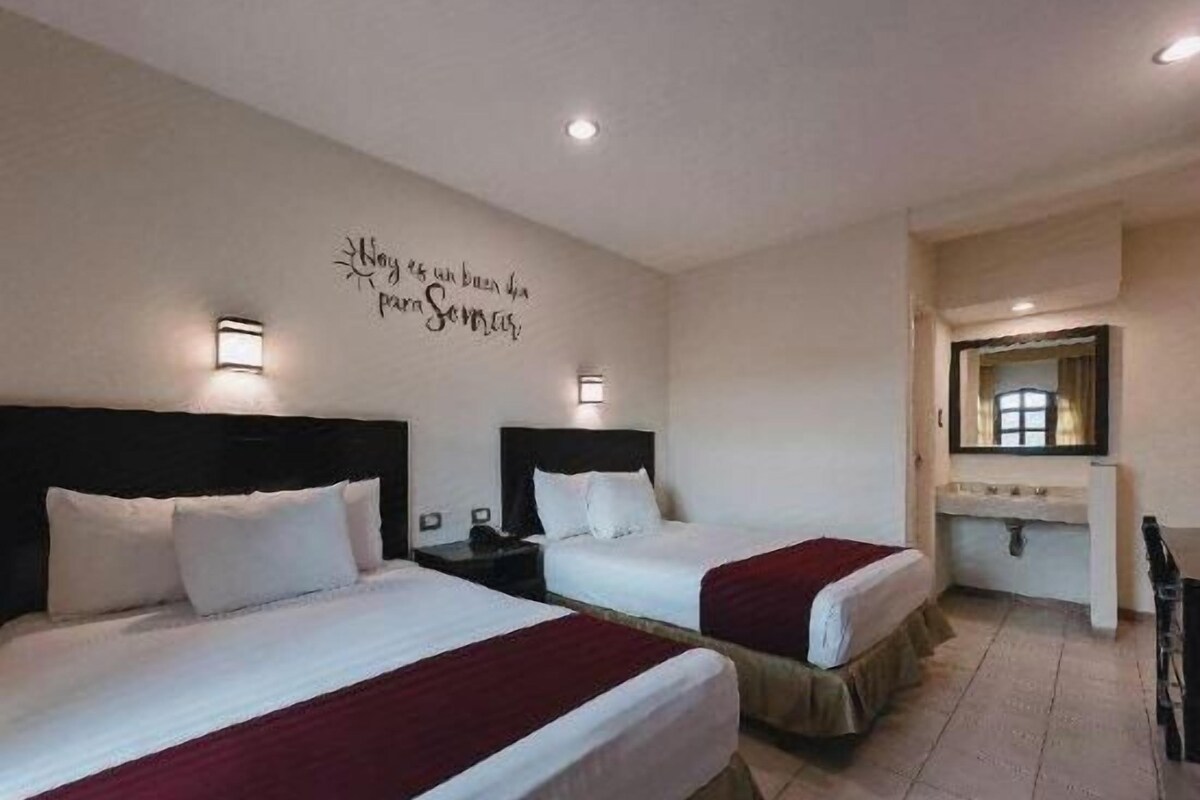 Quarto de hotel com duas camas, banheiro e parede com uma frase motivacional.