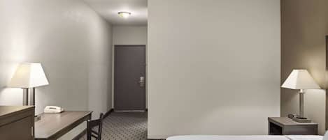 Quarto de hotel com uma cama, uma mesa com telefone, uma cadeira, duas luminárias de cabeceira e uma porta que dá para outro cômodo.