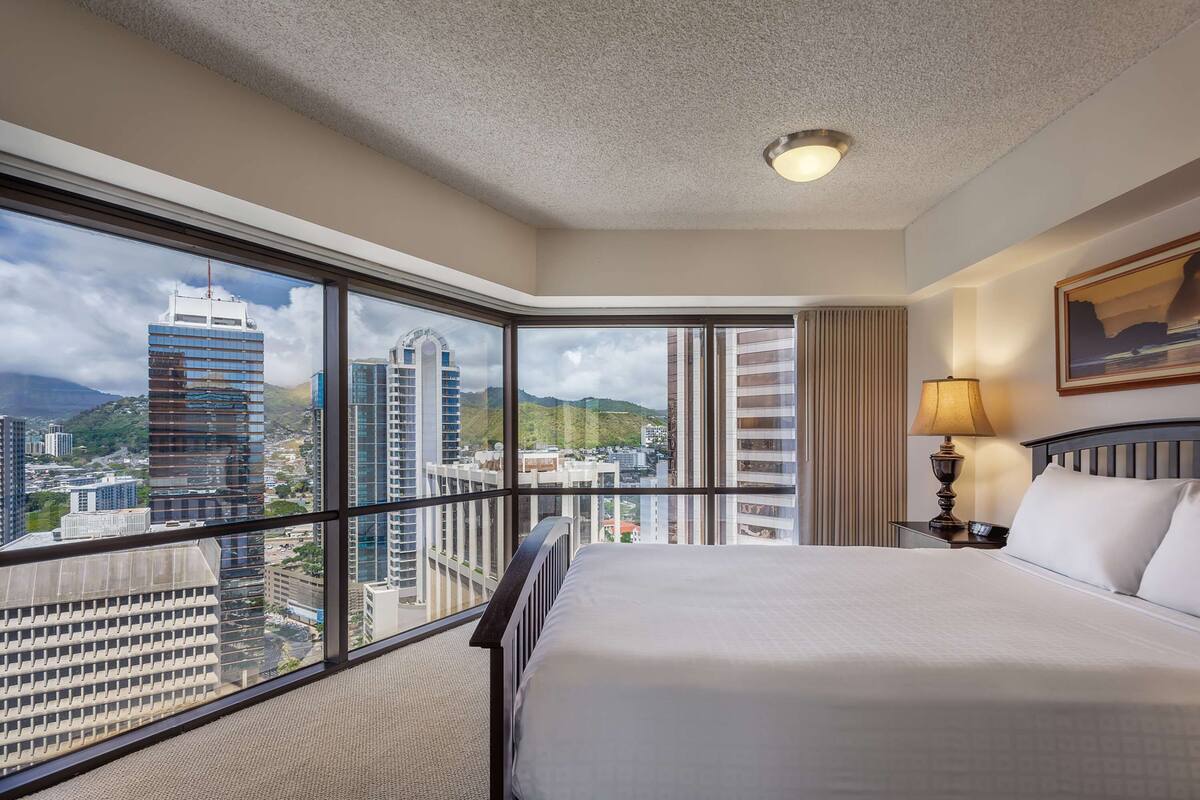 Quarto de hotel com uma grande janela que oferece vista panorâmica da cidade, cama bem arrumada, abajur e quadro na parede.