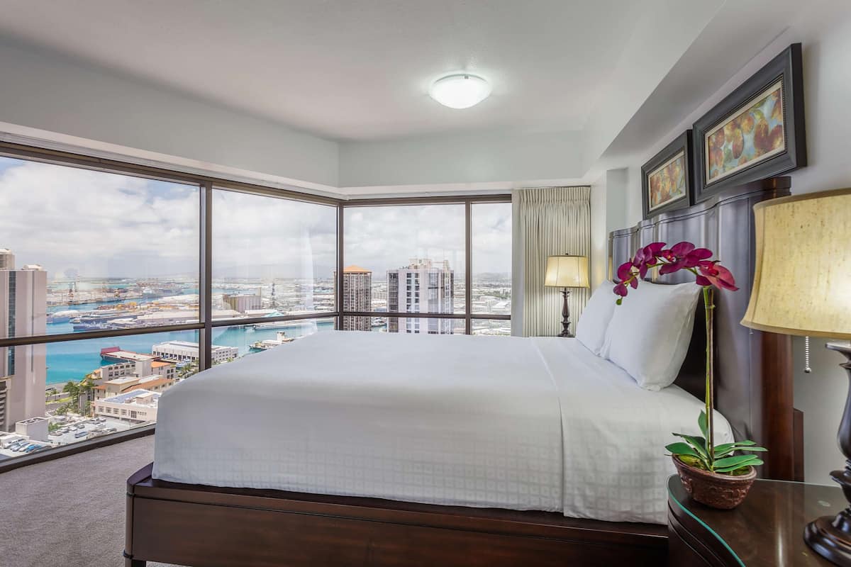 Quarto de hotel com uma cama grande, vista para a paisagem urbana e o porto, uma orquídea em vaso e um abajur.