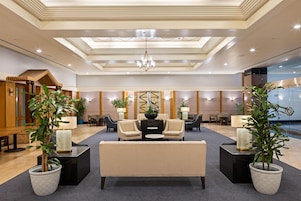 Um saguão de hotel com área de estar central, várias mesas e plantas em vasos.