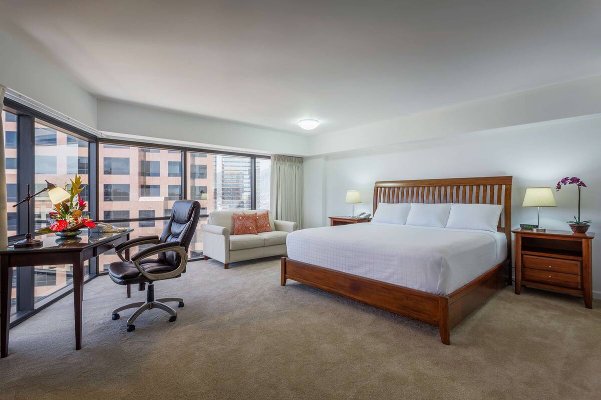 Quarto de hotel com uma cama grande, uma escrivaninha com cadeira, uma mesinha de cabeceira com abajur e vista para a cidade através de grandes janelas.