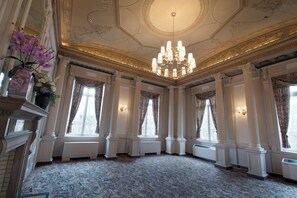 Uma sala imponente com teto ricamente decorado, amplas janelas com cortinas e um lustre.