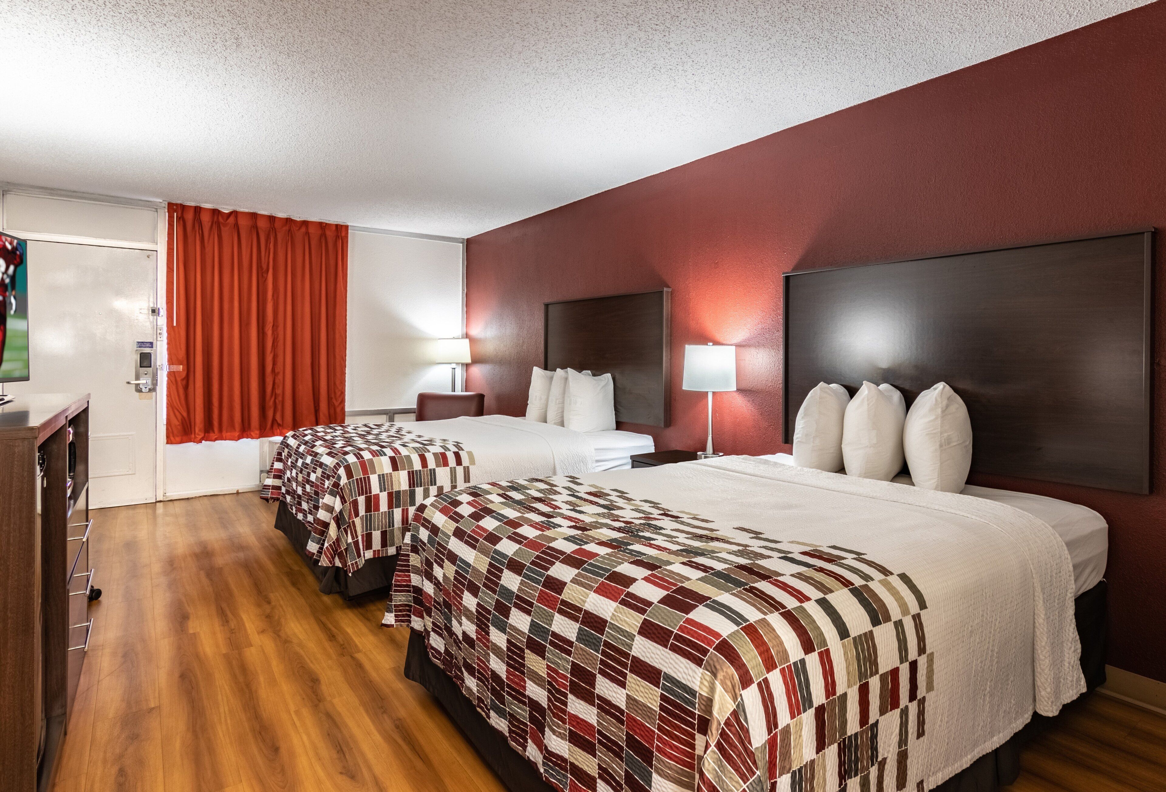 Quarto de hotel com duas camas, piso de madeira, cortina vermelha e televisão na parede esquerda.