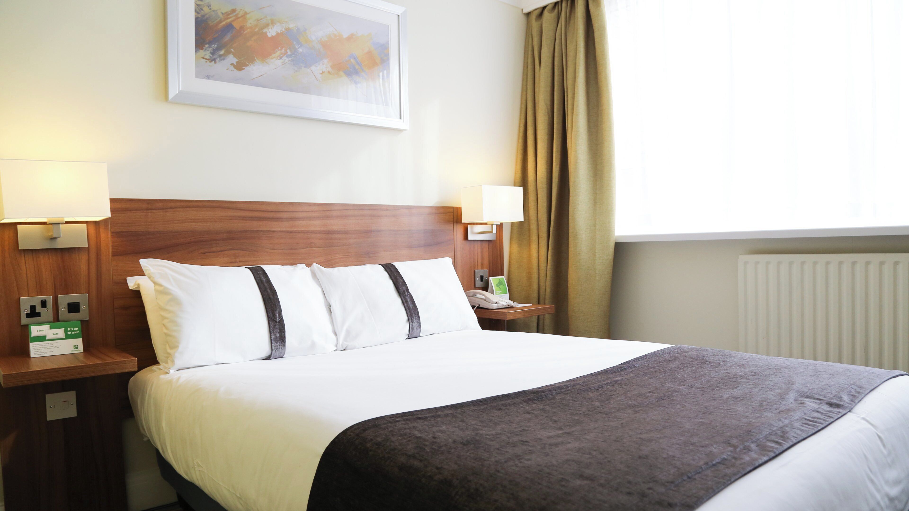 Quarto de hotel com cabeceira de madeira, roupa de cama branca, mesa de cabeceira com abajur, telefone e uma pintura abstrata emoldurada na parede.