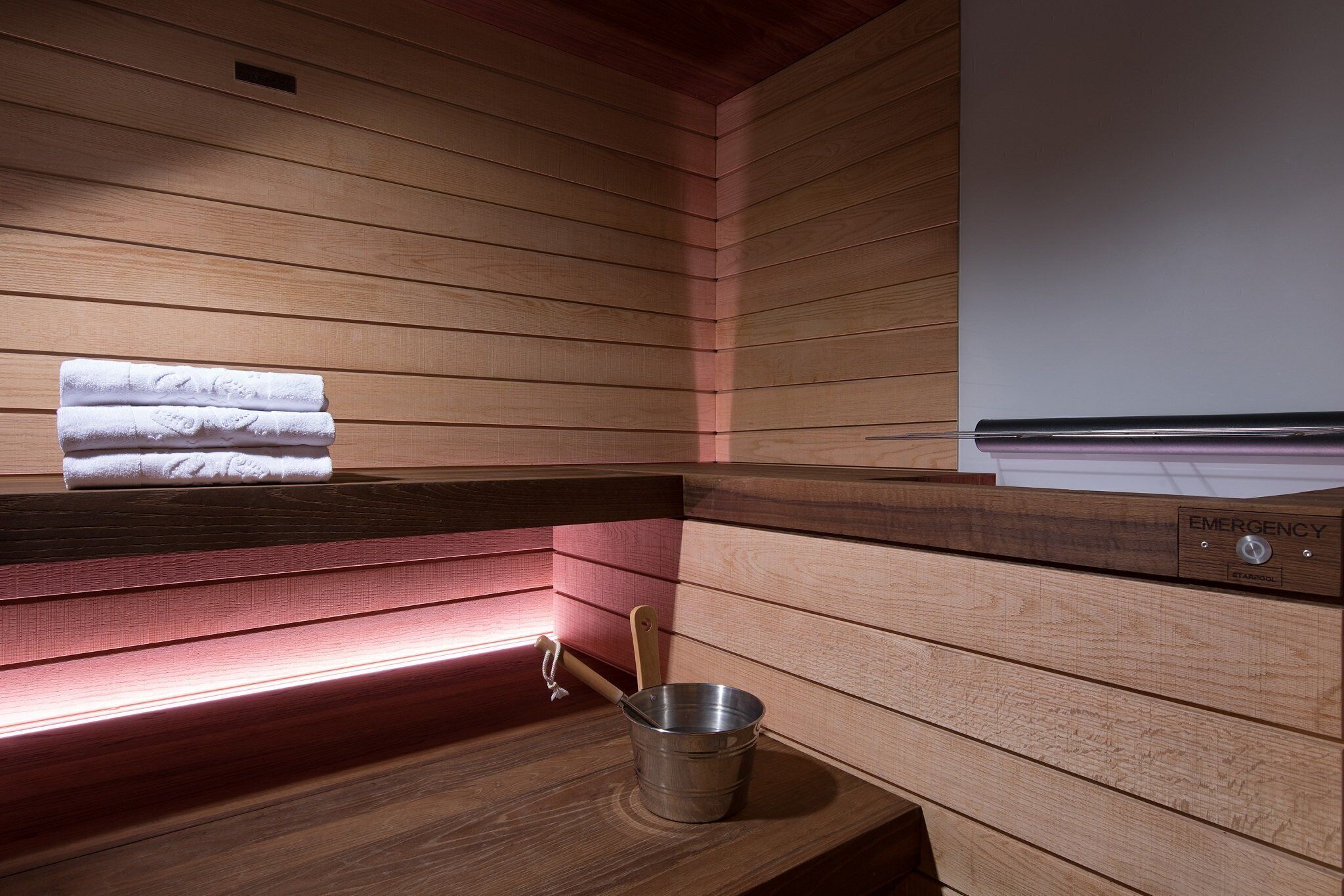 Uma sauna com paredes de madeira, um banco, um porta-toalhas e um balde de metal com cabo de madeira.