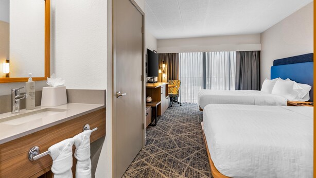 Quarto de hotel com uma cama grande, uma cama menor, uma escrivaninha com cadeira, banheiro com espelho e janela com cortinas.
