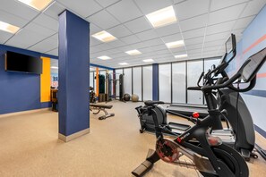 Academia com equipamentos de ginástica, televisão e uma janela grande.
