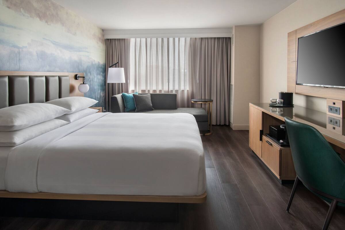 Quarto de hotel com uma cama grande, uma escrivaninha com cadeira, televisão de tela plana e uma janela com cortinas.