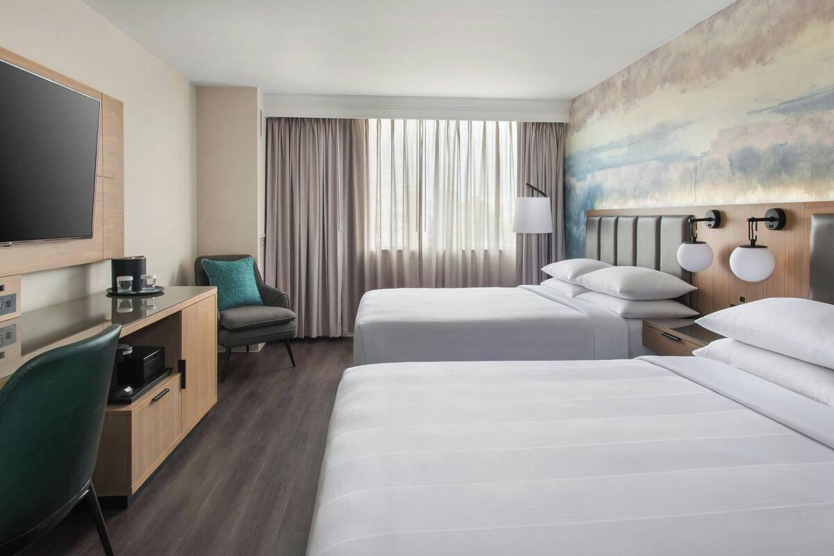 Quarto de hotel com duas camas, uma mesa de trabalho com televisão, uma cadeira e um quadro na parede.