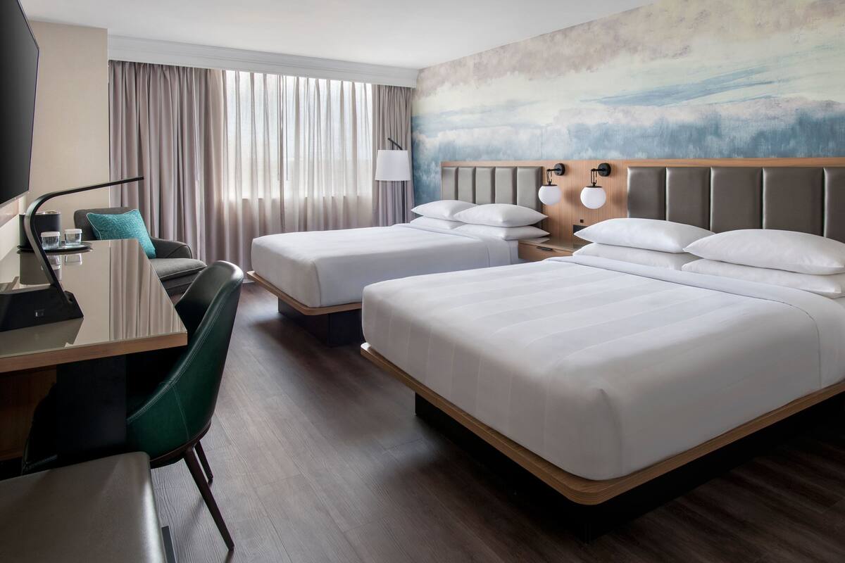 Quarto de hotel com duas camas, uma escrivaninha com cadeira, televisão e um quadro na parede.