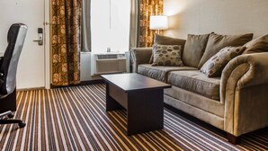 Quarto de hotel com carpete listrado, um sofá com almofadas estampadas, uma mesa e uma cadeira.