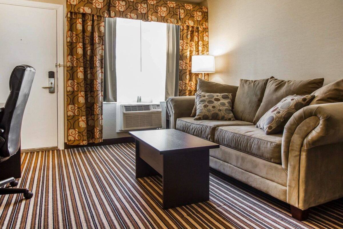 Quarto de hotel com carpete listrado, um sofá com almofadas estampadas, uma mesa e uma cadeira.