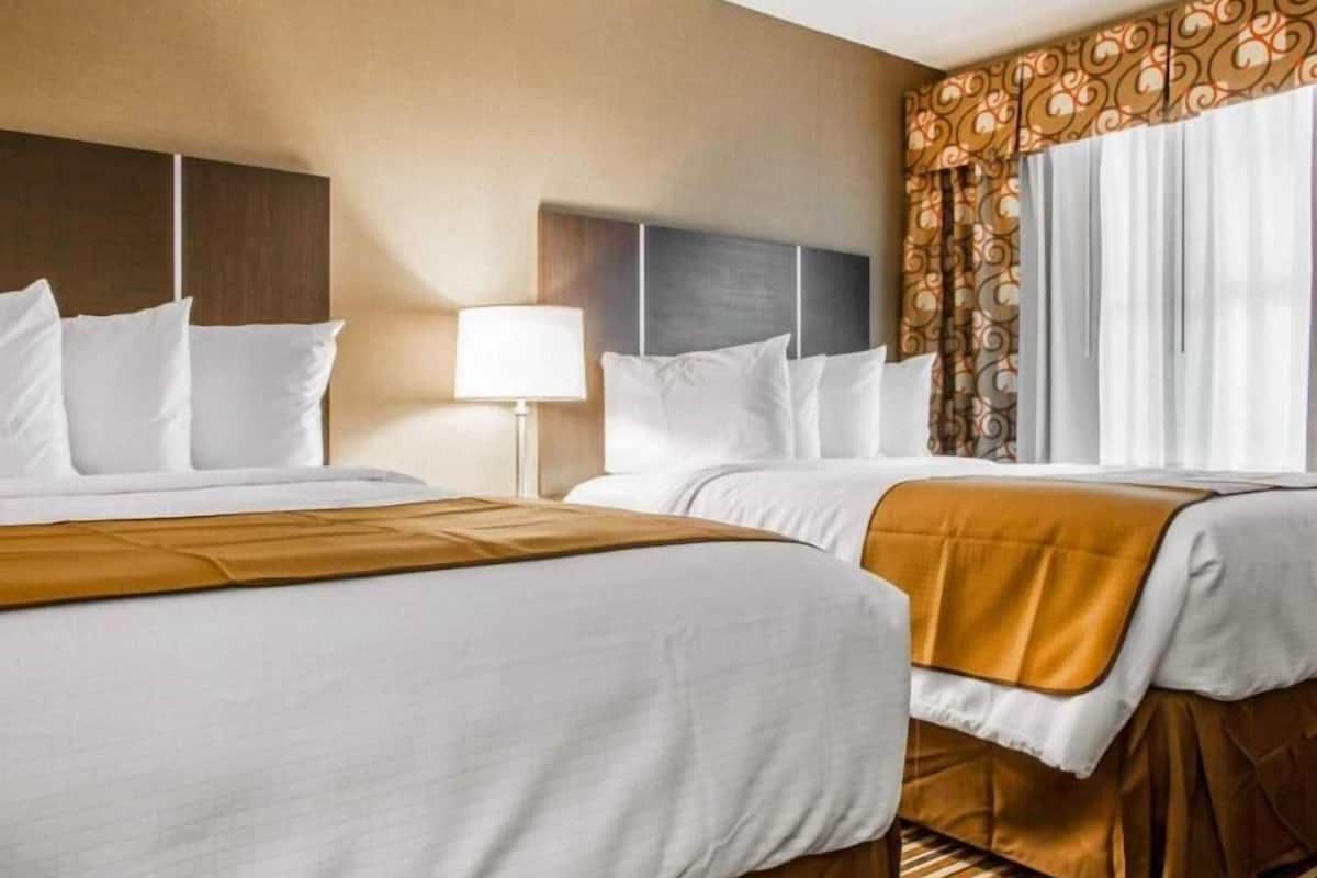 Quarto de hotel com duas camas, uma mesinha de cabeceira com abajur, cortina estampada e uma janela com cortinas brancas.