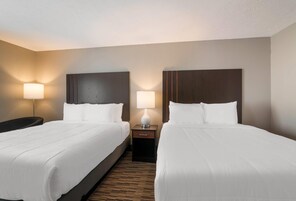 Quarto de hotel com duas camas, cada uma com roupa de cama branca, cabeceira escura e luminária de cabeceira.