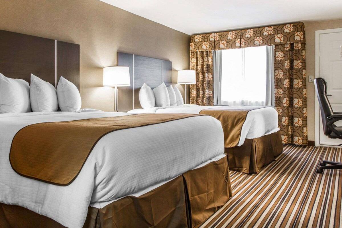 Quarto de hotel com duas camas, uma janela grande com cortinas, uma cadeira e um abajur.