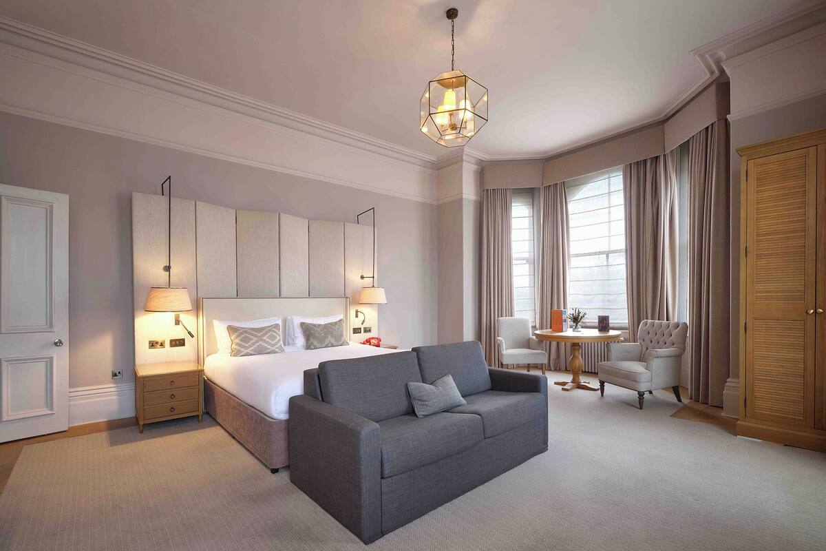 Quarto de hotel com uma cama grande, um sofá cinzento, uma mesa de centro redonda e um guarda-roupa de madeira.