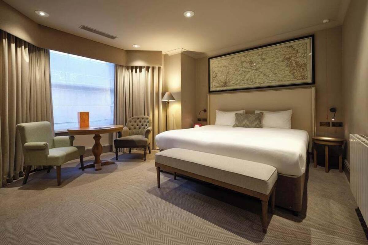 Quarto de hotel com uma cama grande, uma escrivaninha, duas cadeiras, um banco, um mapa na parede e vista da cidade pela janela.