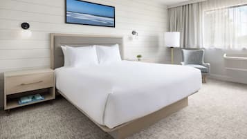 Quarto de hotel com uma cama grande, mesa de cabeceira, uma cadeira, abajur e janela com cortinas.