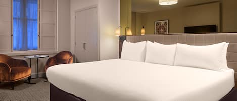 Quarto de hotel com uma cama grande, uma mesa redonda pequena, duas poltronas, uma janela com cortinas azuis e uma televisão na parede.