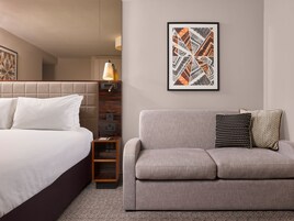 Quarto de hotel com cama, sofá, mesa de cabeceira, luminária e uma obra de arte abstrata emoldurada na parede.