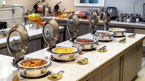 Buffet com diversos alimentos em recipientes fechados, uma cesta de frutas e um canto para café ao fundo.