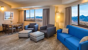 Quarto de hotel com uma grande janela oferecendo vista para as montanhas, um sofá azul, duas poltronas, uma mesa de jantar com cadeiras e um abajur.