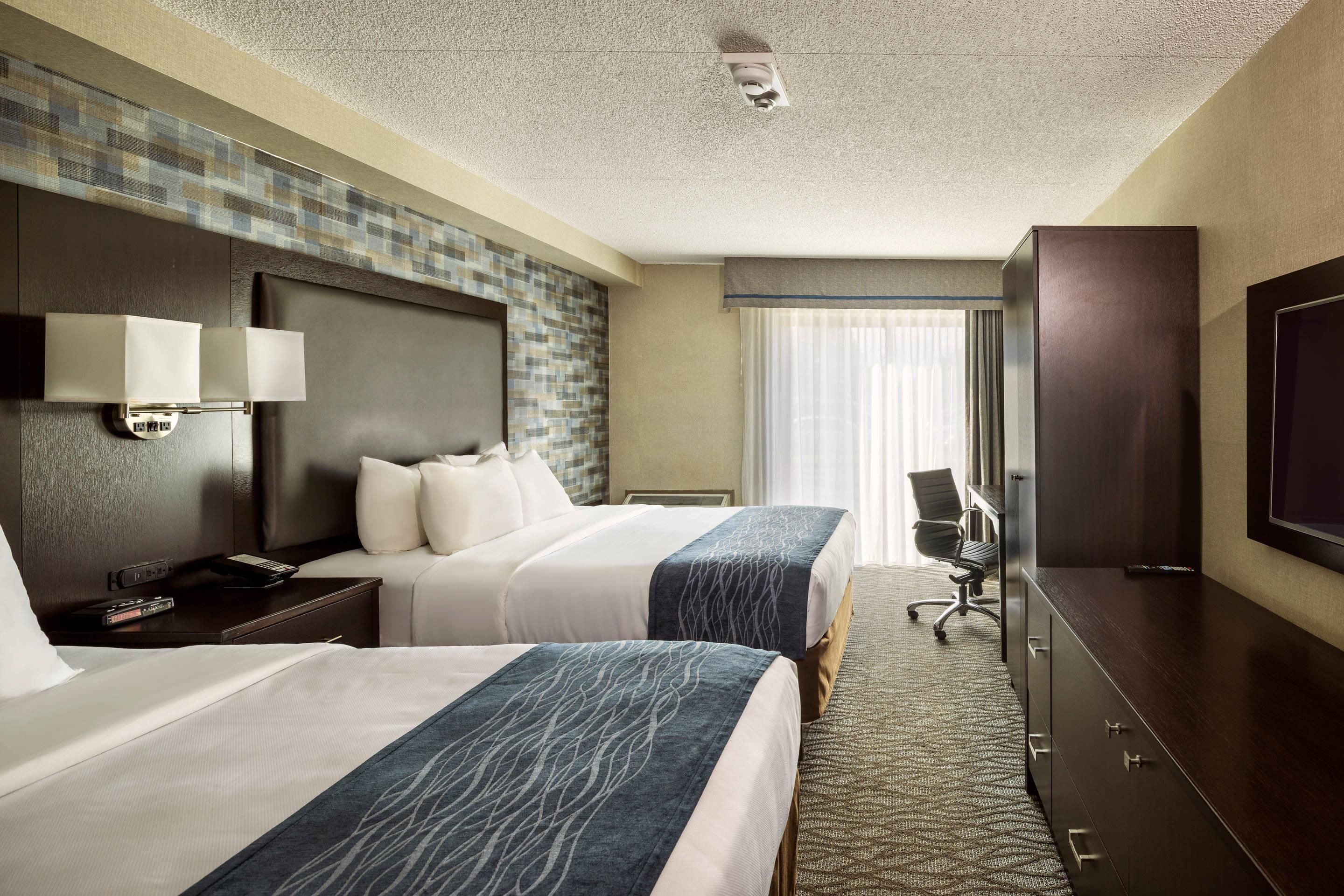 Quarto de hotel com duas camas, uma escrivaninha, uma cadeira, uma televisão e um guarda-roupa.