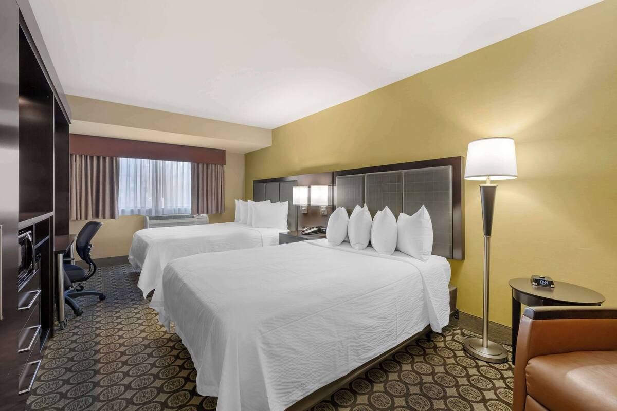 Quarto de hotel com duas camas, uma mesa, uma cadeira, uma luminária e uma janela com cortinas.