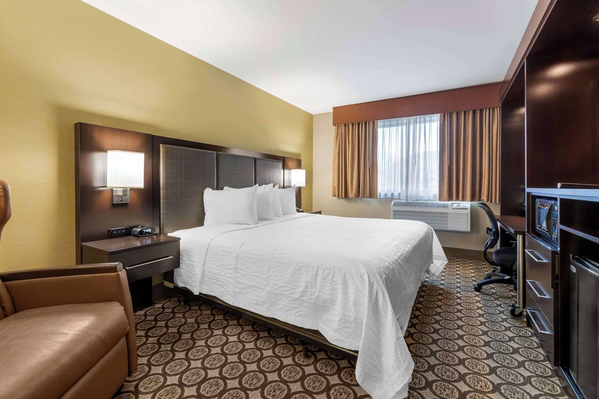 Quarto de hotel com uma cama grande, uma mesa de trabalho com luminária, uma cadeira, um minibar e uma janela ampla com cortinas.