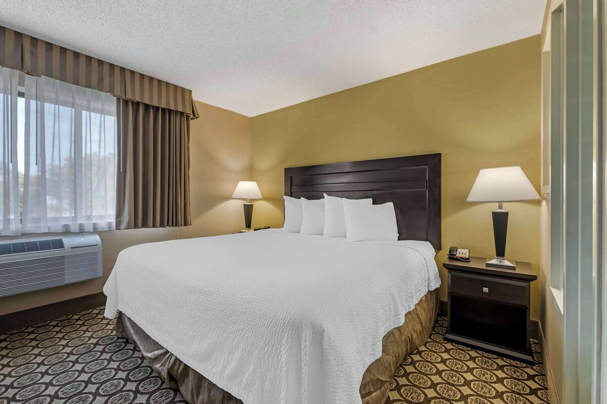 Quarto de hotel com uma cama grande, duas luminárias de cabeceira, uma mesa de apoio e uma janela com cortinas.