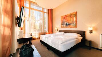 Quarto de hotel com cama, escrivaninha, cadeira, televisão e um quadro na parede.
