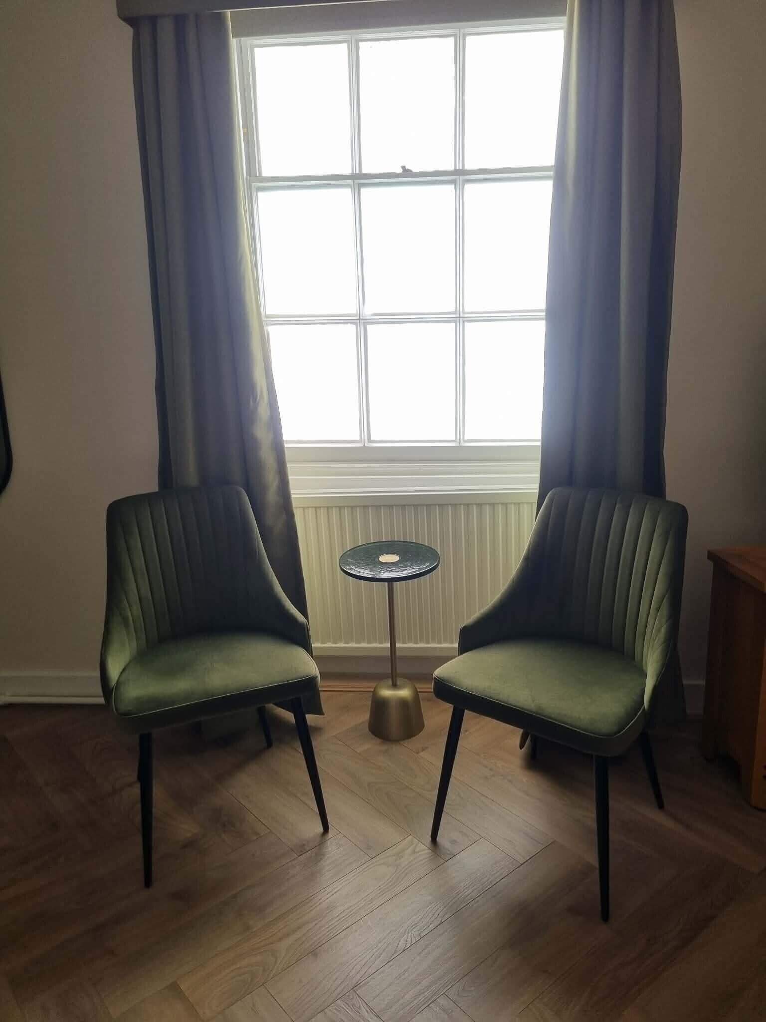 Um quarto com duas poltronas verdes, uma mesa redonda pequena e uma janela com cortinas.
