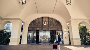 Entrada de um hotel com uma imponente arcada, uma placa com a inscrição "BRICKSTONE'S" e uma pessoa puxando uma mala.