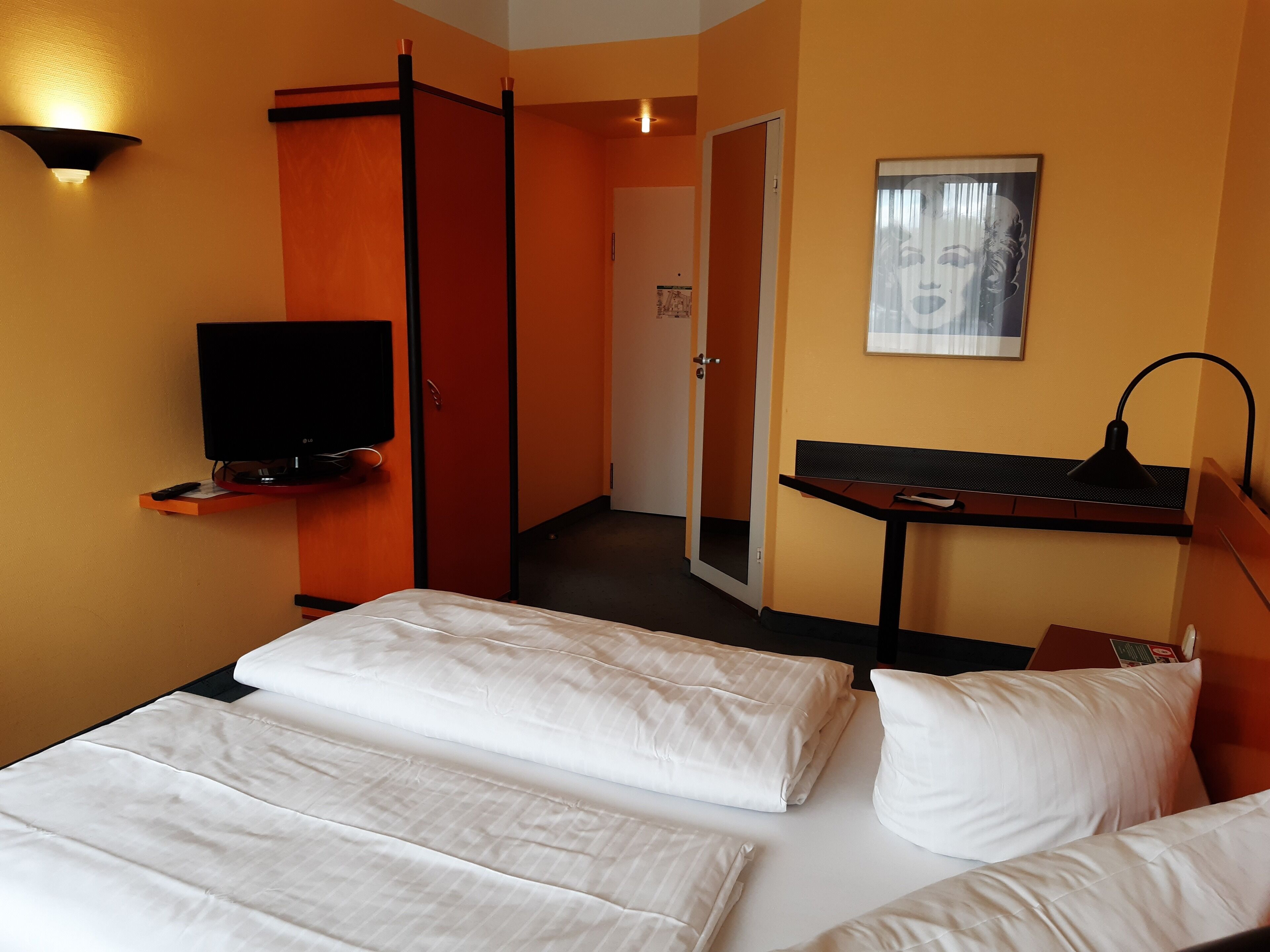 Quarto de hotel com cama, televisão, escrivaninha e porta.
