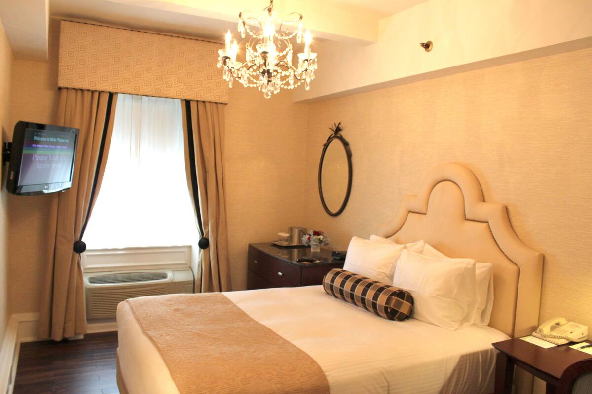 Quarto de hotel com cama, televisão na parede, escrivaninha com telefone e janela com cortinas.